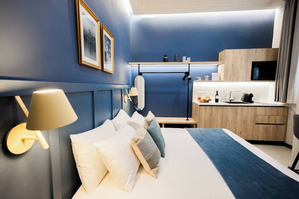 Quarto de hotel com cama, luminárias de cabeceira, escrivaninha com espelho, pequeno espaço de cozinha e televisão.