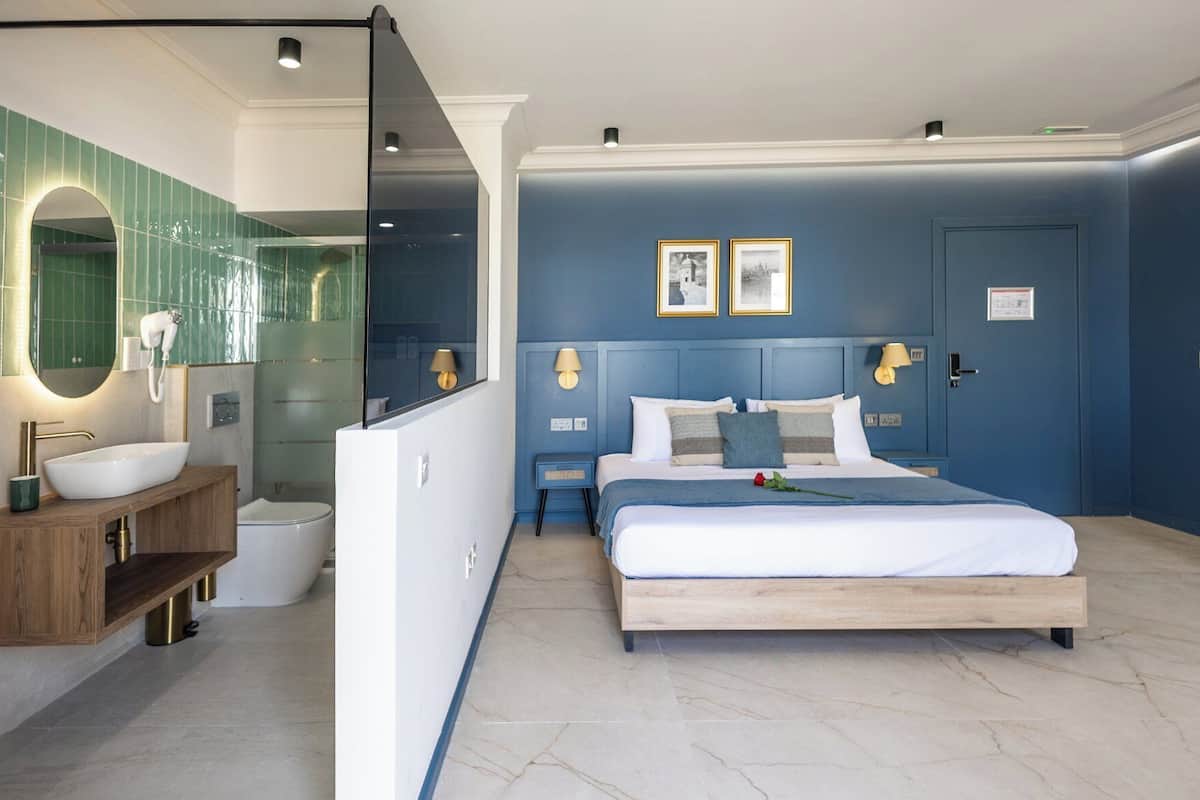 Quarto moderno com uma cama grande, mesa de cabeceira, espelho, toucador com pia e área de chuveiro.