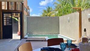 Uma área de refeições ao ar livre com uma mesa posta para dois, piscina ao fundo e vista para palmeiras.