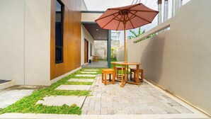 Um corredor exterior estreito com uma mesa de madeira e cadeiras, um guarda-sol vermelho e um caminho de tijolos com grama.
