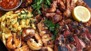 Tábua de madeira com carnes grelhadas, camarão e batatas fritas, acompanhada de molhos para servir.