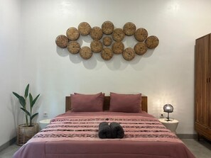 Quarto com cama, almofadas, mesa de cabeceira com abajur, objeto decorativo na parede e planta em vaso.