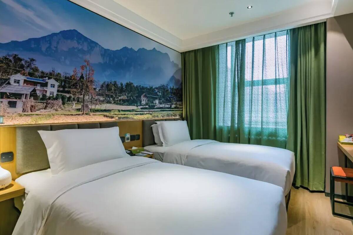 Quarto de hotel com duas camas, uma janela grande com cortinas verdes e um mural de uma paisagem montanhosa.