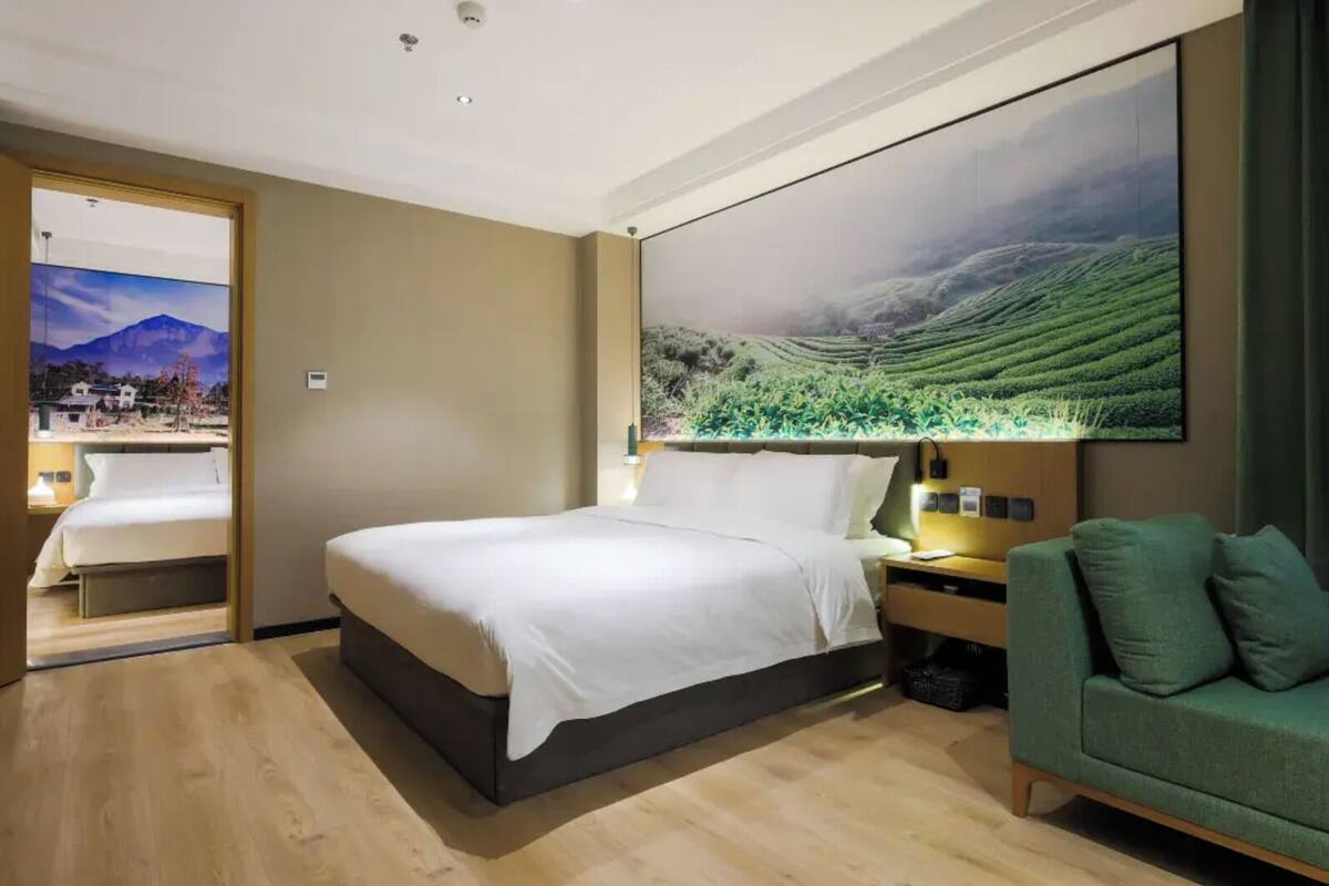 Quarto de hotel com uma cama grande, um sofá verde, uma mesa de cabeceira e uma televisão fixada na parede.