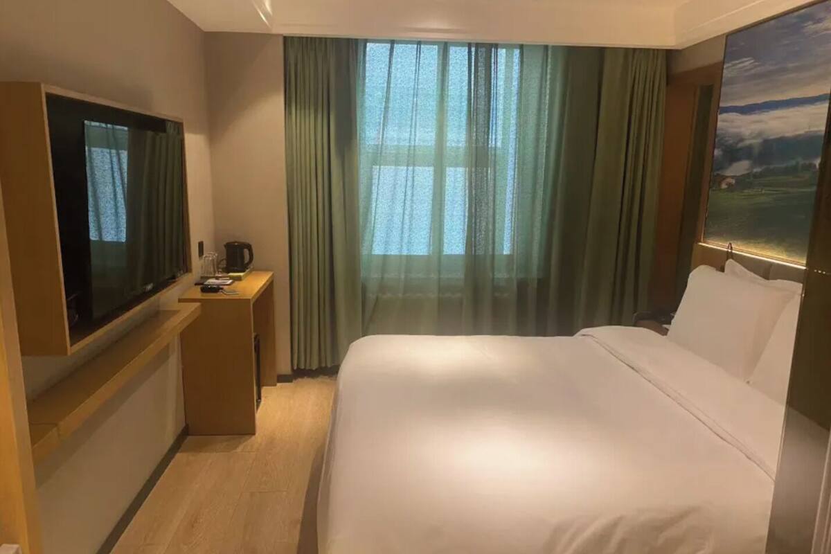 Quarto de hotel com uma cama grande, uma televisão de tela plana, uma pequena mesa com cafeteira e uma janela com cortinas.