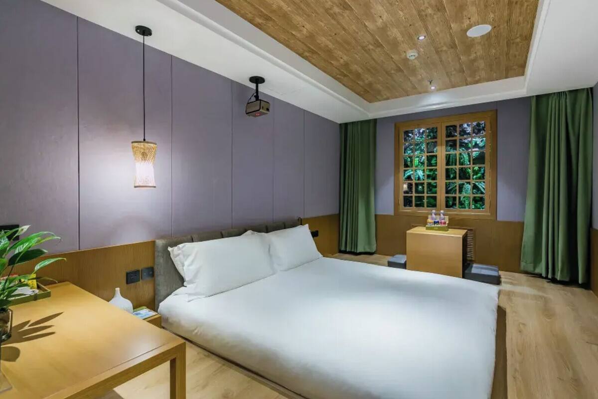 Quarto de hotel com uma cama grande, cabeceira de madeira, mesa de cabeceira com abajur, pequena escrivaninha e janela com cortinas.