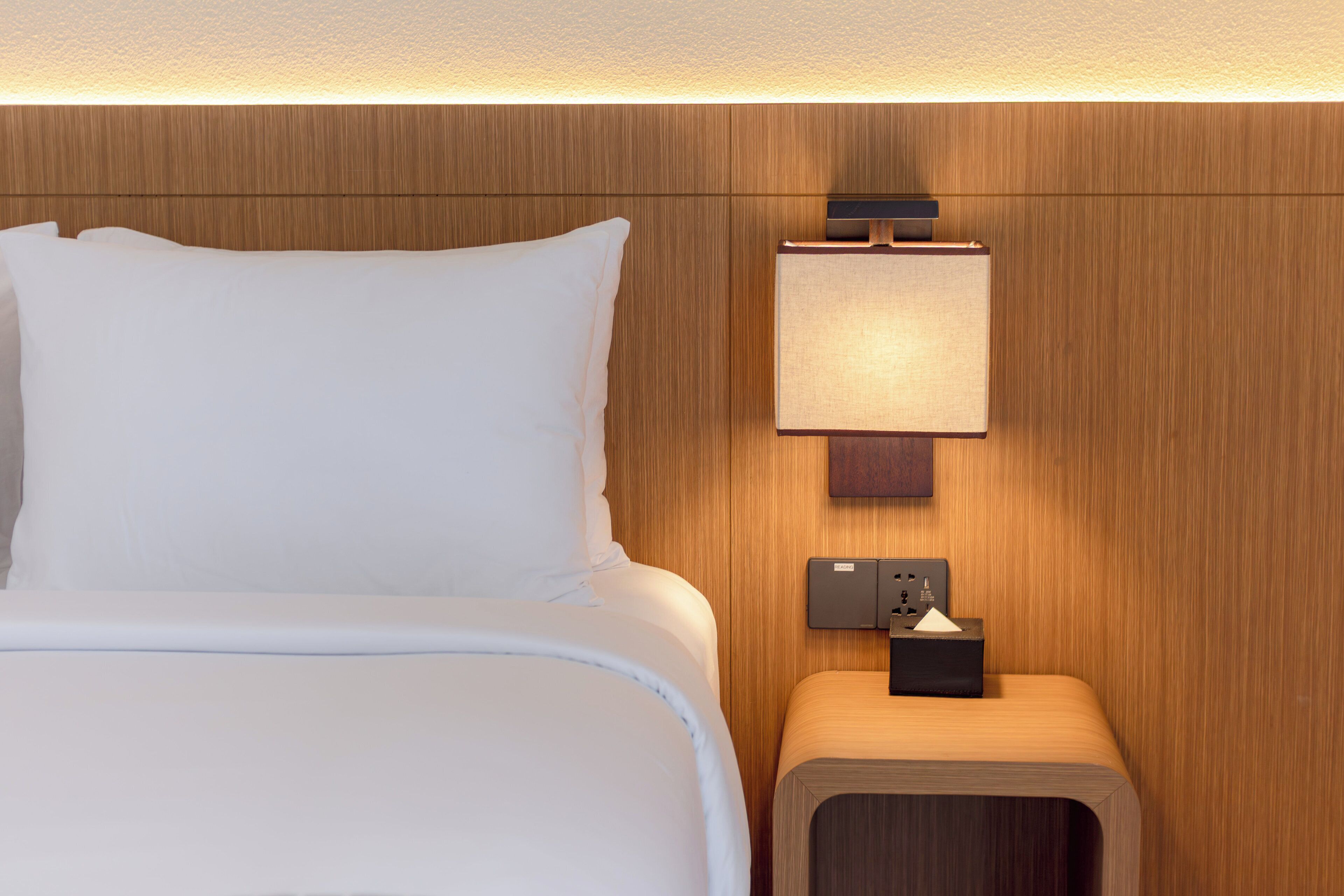 Uma cama com roupa de cama branca, um criado-mudo com abajur e uma tomada.