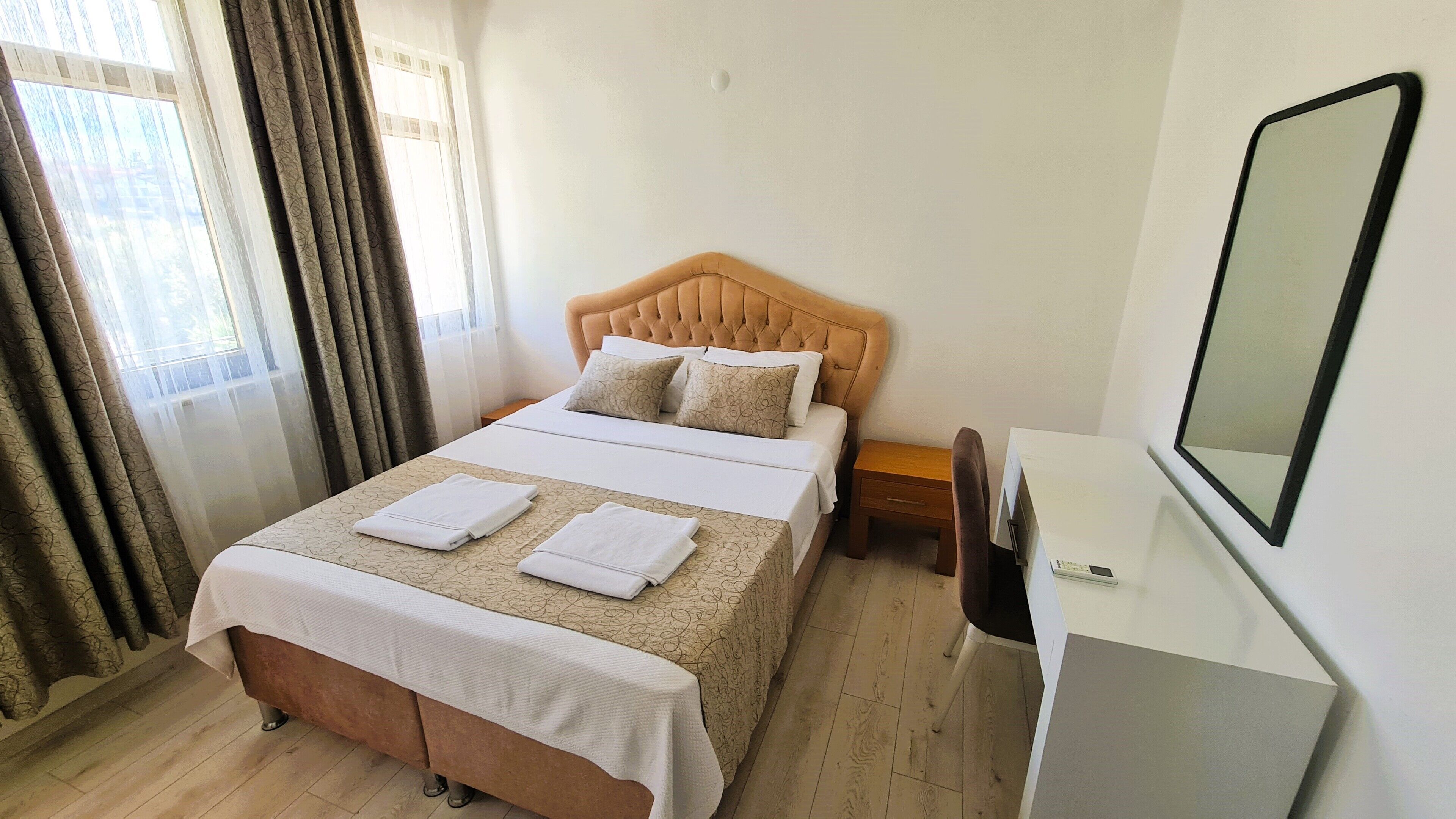 Uma cama bem arrumada com cabeceira, mesa de cabeceira, espelho e janela com cortinas.