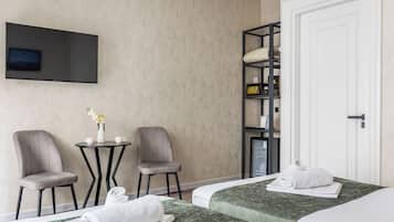 Quarto de hotel com duas camas, uma mesa pequena com cadeiras, uma televisão fixada na parede e uma estante com diversos objetos.