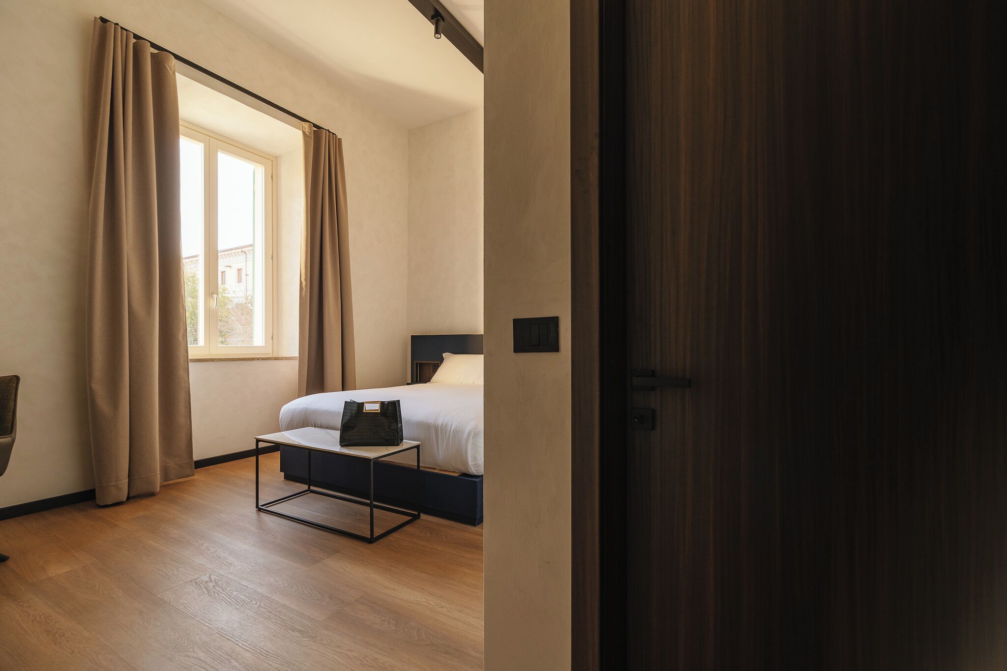 Quarto de hotel moderno com uma cama grande, uma mesinha de cabeceira e vista para o exterior.