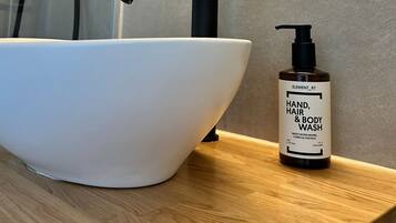 Casa de banho com lavatório suspenso branco, torneira preta e frasco de gel de banho, cabelo e corpo sobre bancada de madeira.