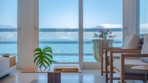 Quarto com uma grande janela com vista para a água, um sofá, uma mesa de centro de madeira com um livro e uma planta em vaso.