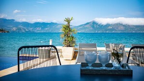 Mesa com cadeiras à beira da piscina, um vaso de planta e um copo de água, com vista para o mar azul e montanhas.