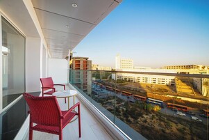 Uma varanda com cadeiras de vime vermelhas e uma pequena mesa, com vista para um horizonte urbano com edifícios e uma rua movimentada com ônibus.