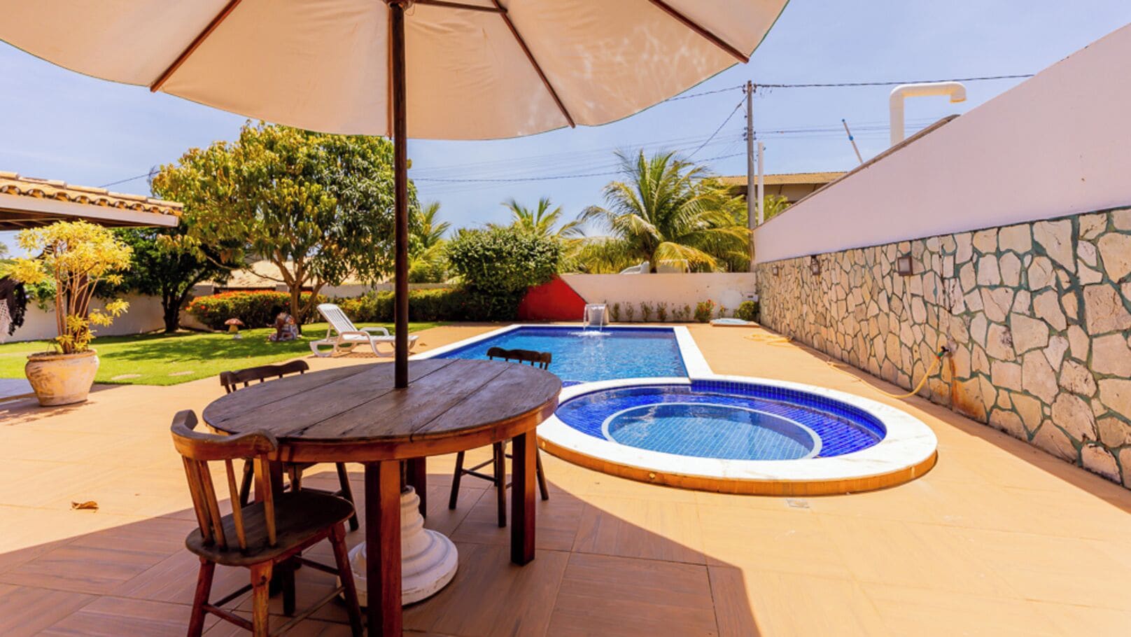 Uma área de piscina com uma mesa e cadeiras de madeira, uma parede de pedra e um objeto esférico vermelho.