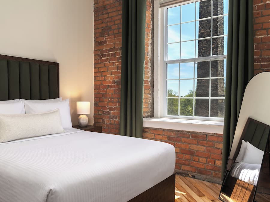 Quarto de hotel com cama, mesa de cabeceira, luminária, janela com vista para um edifício de tijolos e espelho.