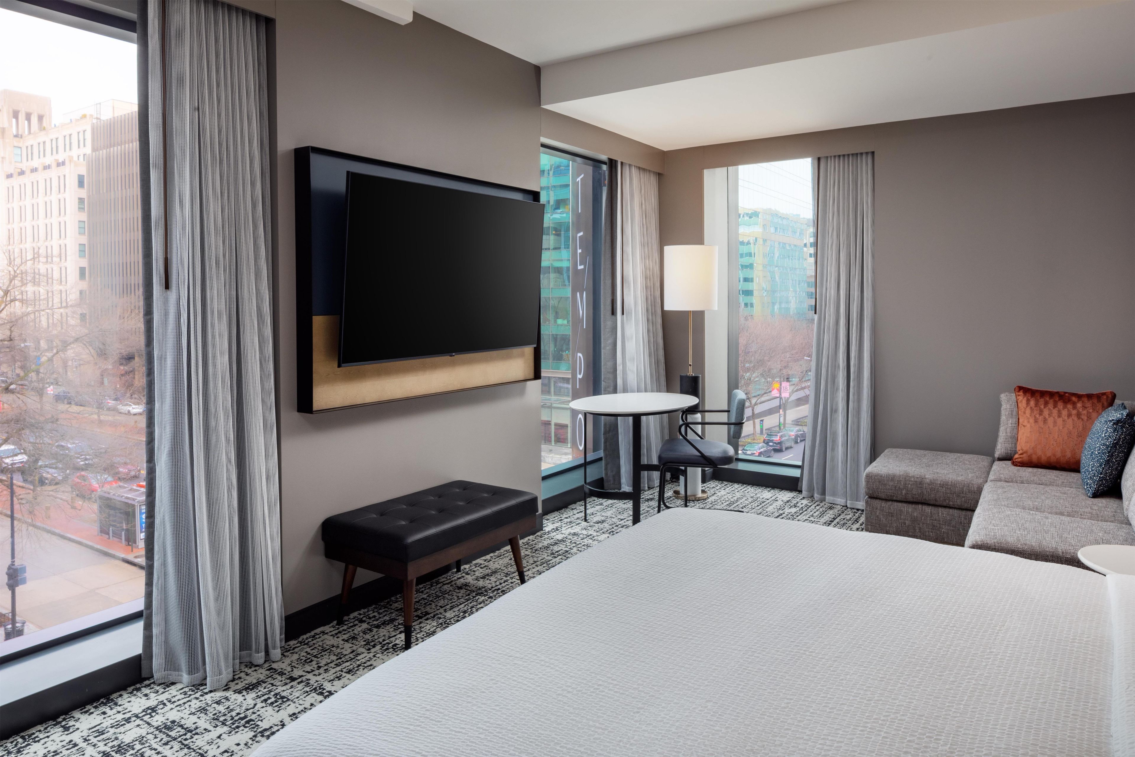 Quarto de hotel com uma cama grande, televisão de tela plana na parede, mesa redonda pequena com uma cadeira e um sofá com almofadas.