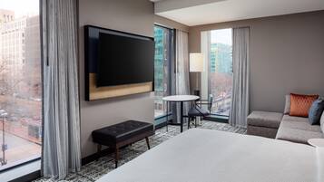 Quarto de hotel com uma cama grande, televisão de tela plana na parede, mesa redonda pequena com uma cadeira e um sofá com almofadas.