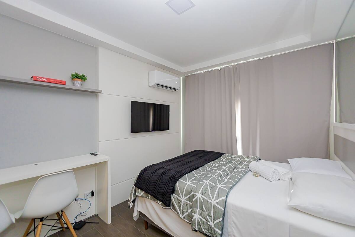 Quarto moderno com cama, escrivaninha, cadeira, televisão e planta em vaso.