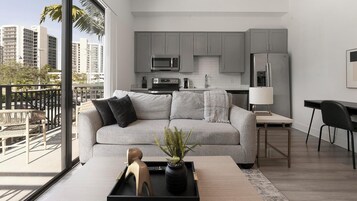 Apartamento moderno com sofá cinza, área de refeições, cozinha com armários cinza e vista para palmeiras e edifícios.