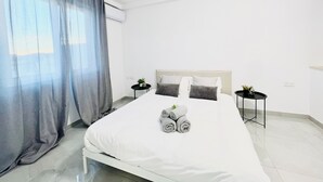 Quarto de hotel moderno com uma cama grande, duas mesas de cabeceira, uma pequena planta em vaso e uma janela com cortinas.