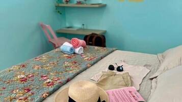 Quarto com cama, chapéu, bolsa rosa e óculos de sol sobre a cama. Há uma janela com persianas e uma estante com uma planta.