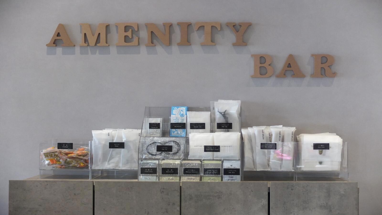Exposição de comodidades no Amenity Bar, incluindo diversos produtos embalados e uma caixa de plástico transparente com itens.