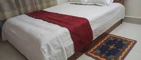 Uma cama bem arrumada com roupa de cama branca e um cobertor vermelho, um tapete estampado no chão e uma cortina parcialmente aberta.