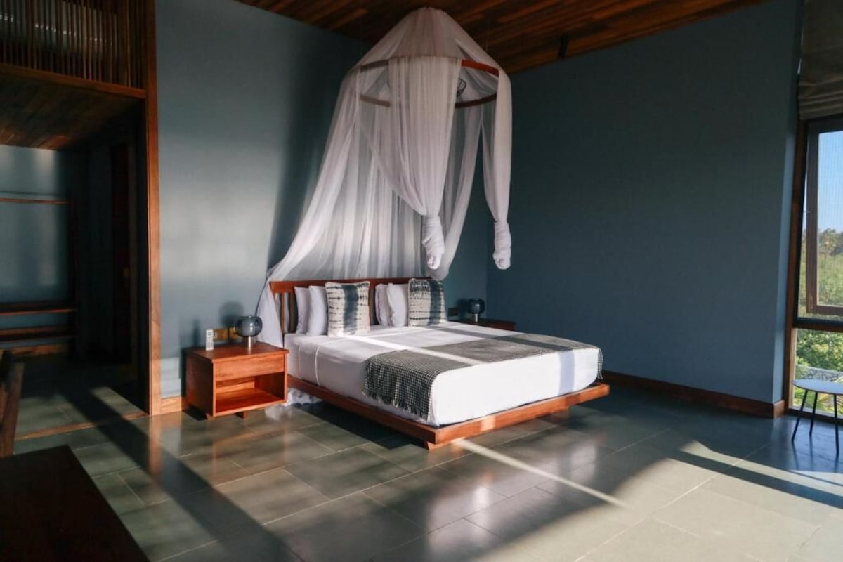 Quarto com cama de dossel, cabeceira de madeira, mesas de cabeceira e vista para o verde lá fora.