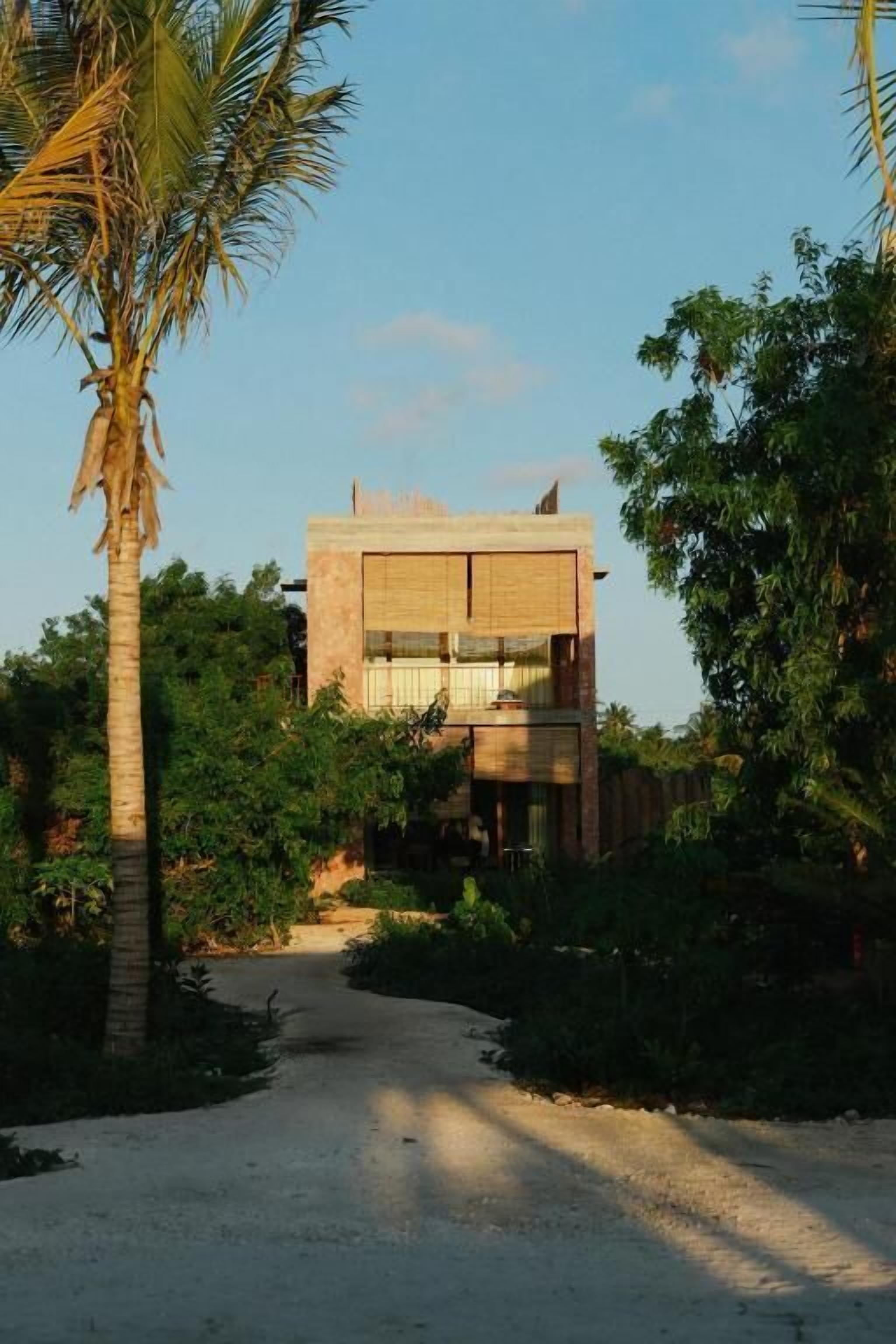 Um edifício moderno com teto plano e janelas amplas, rodeado por vegetação exuberante e uma trilha de terra.