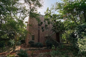 Um edifício rústico de pedra rodeado por vegetação densa, com um caminho bem definido a levar à entrada.