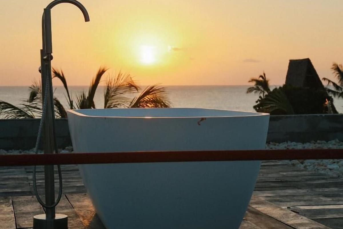 Uma banheira branca em um deck de madeira com vista para o pôr do sol sobre o oceano.