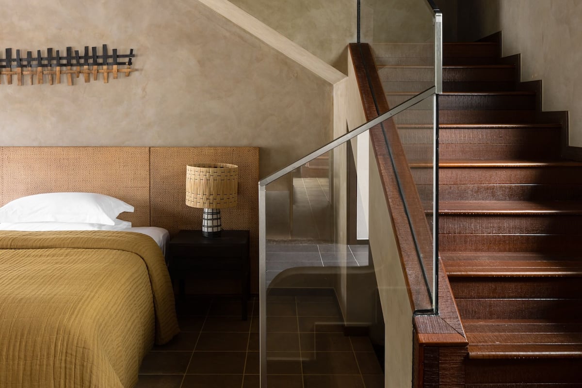 Quarto de hotel moderno com escada de madeira, cama com manta bege, mesa de cabeceira com abajur e luminária na parede.