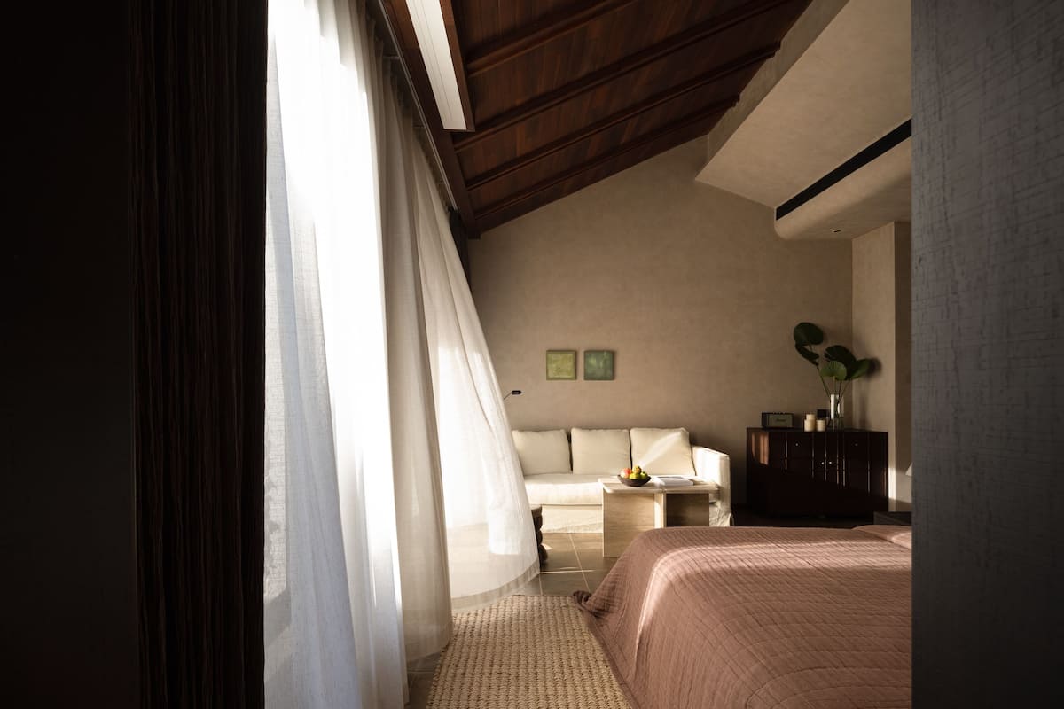 Quarto com cama, mesa de cabeceira, sofá e janela com cortinas finas.