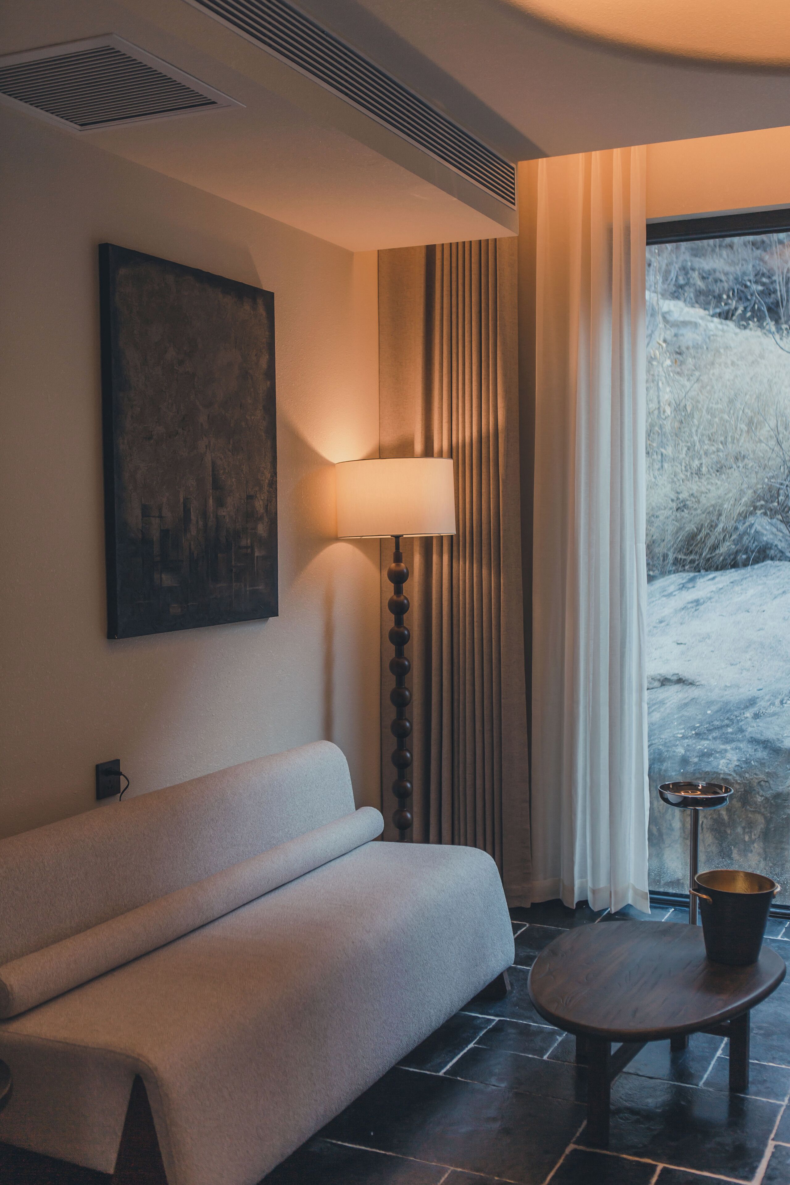 Quarto de hotel moderno com sofá, luminária, mesa pequena e vista para uma paisagem nevada pela janela.