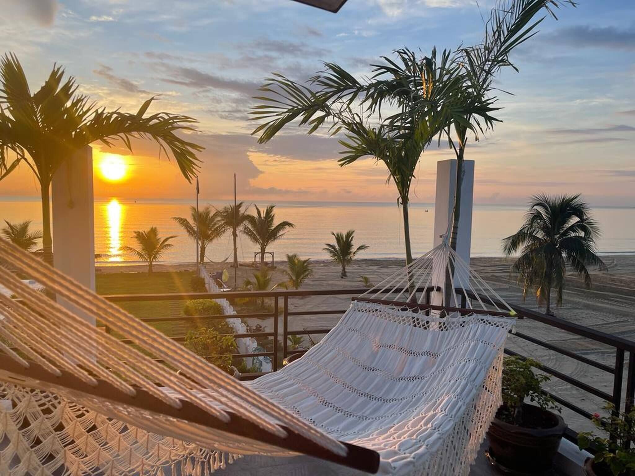 Uma rede numa varanda com vista para uma praia, palmeiras e um pôr do sol.