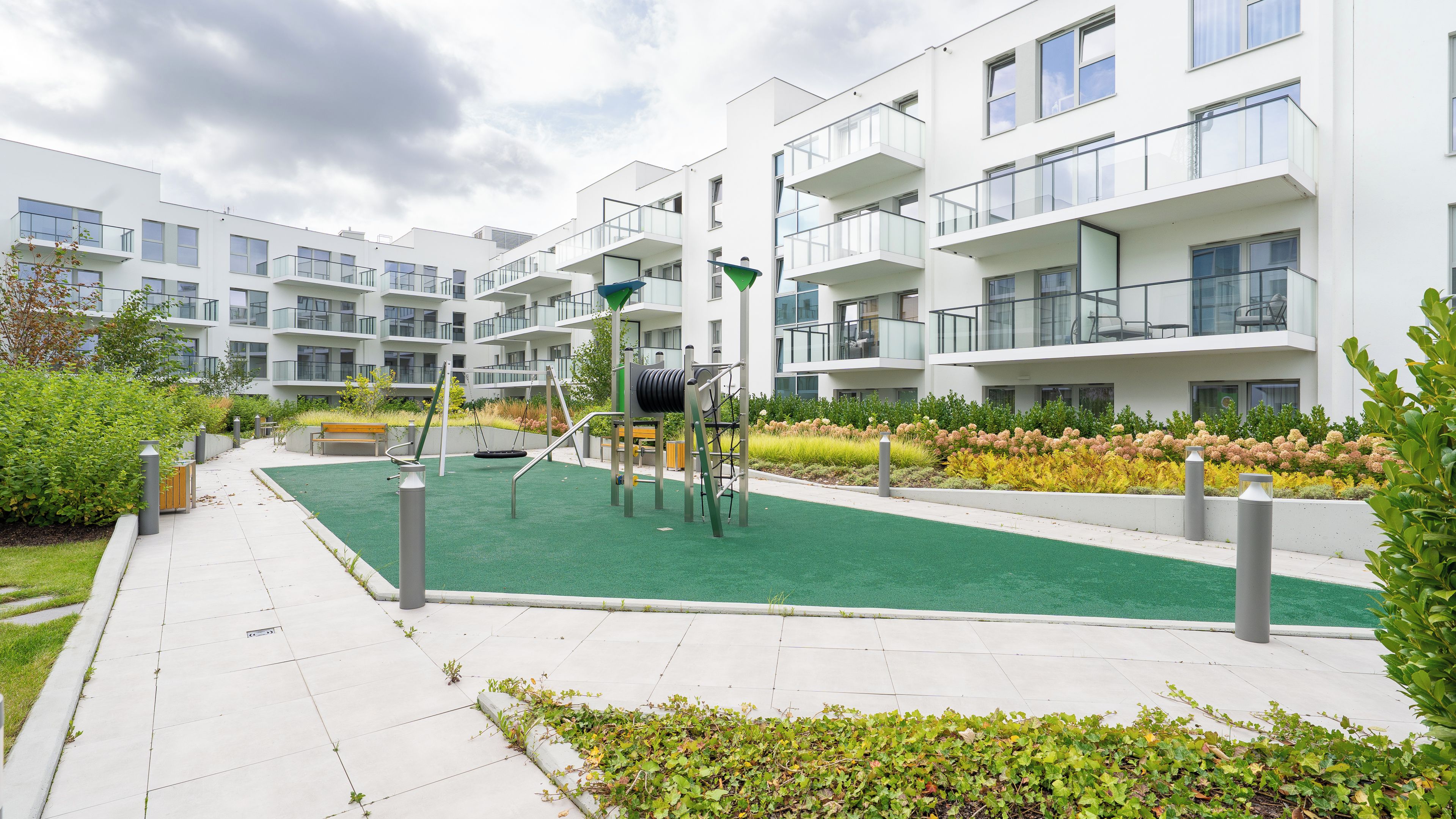 Um moderno conjunto de apartamentos com área de recreação infantil, incluindo balanços e escorrega, rodeado por áreas verdes bem cuidadas.