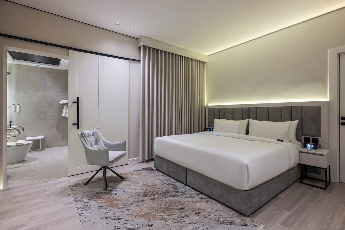 Quarto moderno com uma cama grande, mesa de cabeceira com relógio, uma cadeira e um banheiro visível por uma porta aberta.
