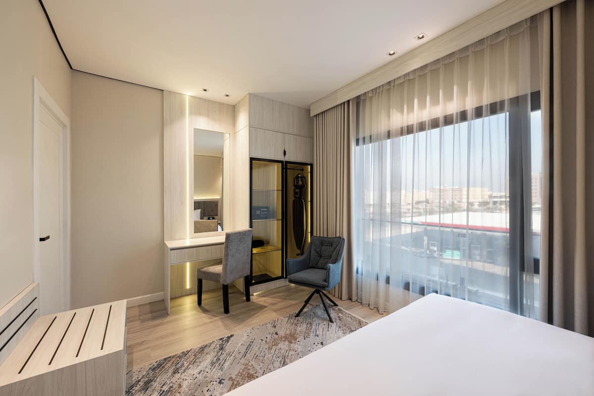 Quarto de hotel moderno com uma cama grande, uma escrivaninha com cadeira, um guarda-roupa e uma janela ampla com cortinas.
