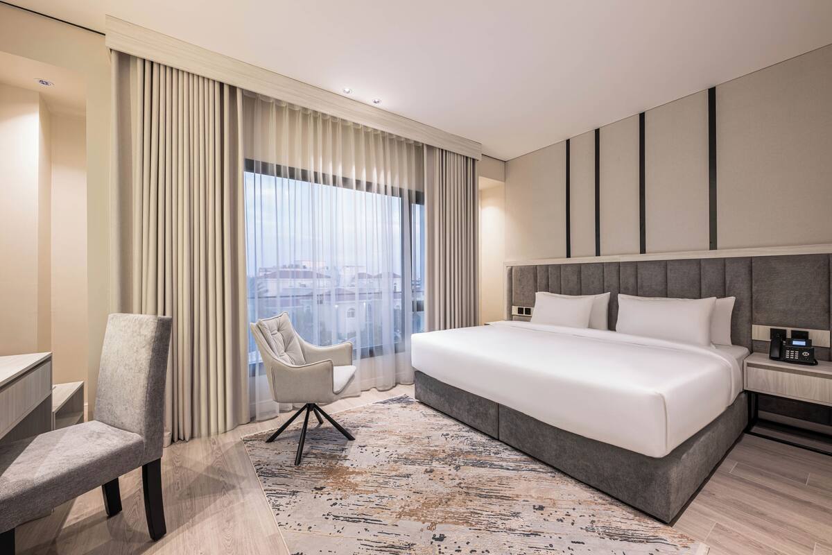 Quarto de hotel moderno com uma cama grande, uma escrivaninha, uma cadeira e vista da cidade através de amplas janelas.