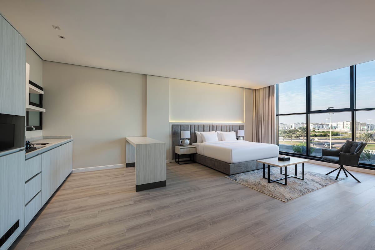 Quarto de hotel moderno com uma cama grande, uma escrivaninha, uma cadeira e vista para a cidade através de amplas janelas.