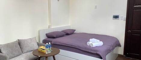 Quarto de hotel bem arrumado com cama, sofá cinza, mesa redonda pequena com artigos de higiene e piso de madeira.