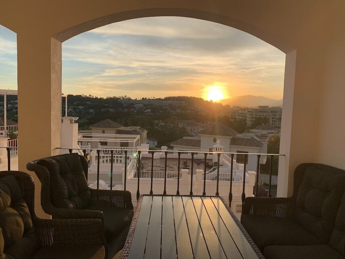 Varanda com móveis de vime e vista para o pôr do sol sobre um bairro residencial.