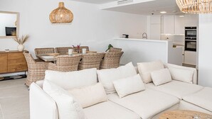 Uma sala de estar moderna com sofá bege, conjunto de jantar em vime e uma cozinha ao fundo.