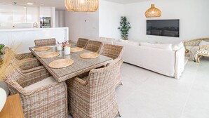 Cozinha moderna com armários brancos, sala de jantar com mobiliário de vime e sala de estar com sofá branco e televisão.