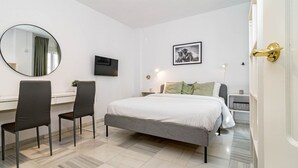 Quarto moderno com uma cama, duas cadeiras, uma escrivaninha, um espelho, uma televisão e um quadro pendurado na parede.