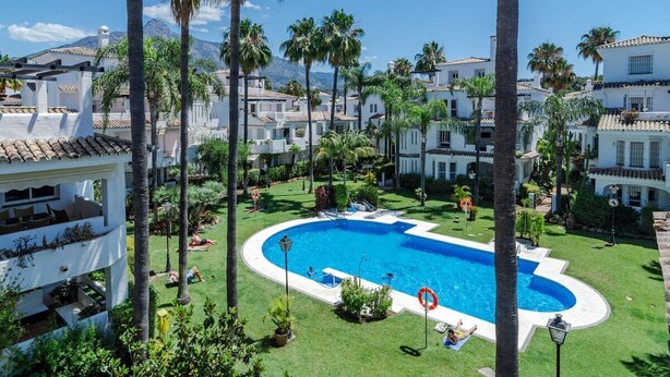 Piscina rodeada por palmeiras e edifícios brancos, com montanhas ao fundo.
