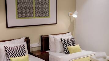Quarto de hotel com duas camas de solteiro, cada uma com colcha verde e roupa de cama branca. Há uma obra de arte emoldurada acima das camas e uma luminária na parede à direita.