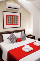 Cama bem arrumada com lençóis brancos, almofadas estampadas em vermelho e preto e manta vermelha.