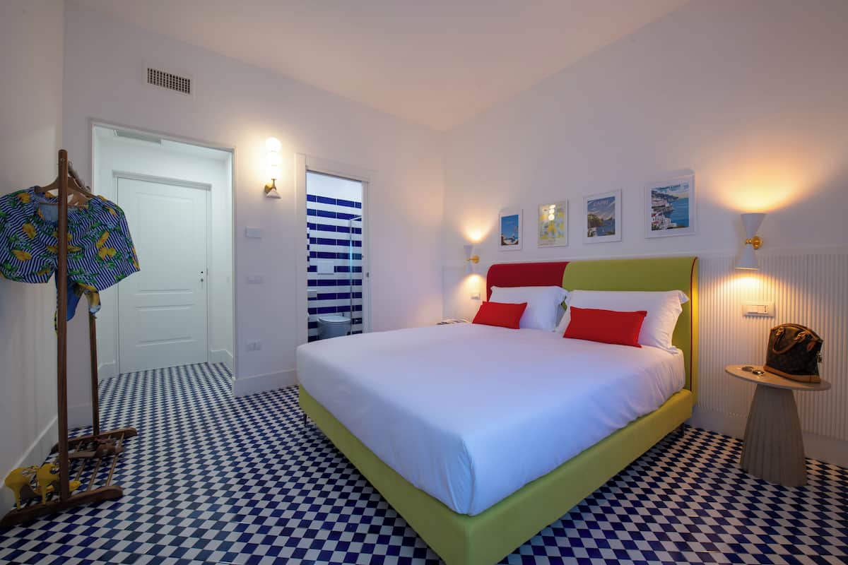 Quarto de hotel com piso xadrez, cama com roupa de cama branca e vermelha, uma cadeira de madeira, uma mesa pequena com abajur e uma porta com cortina listrada.